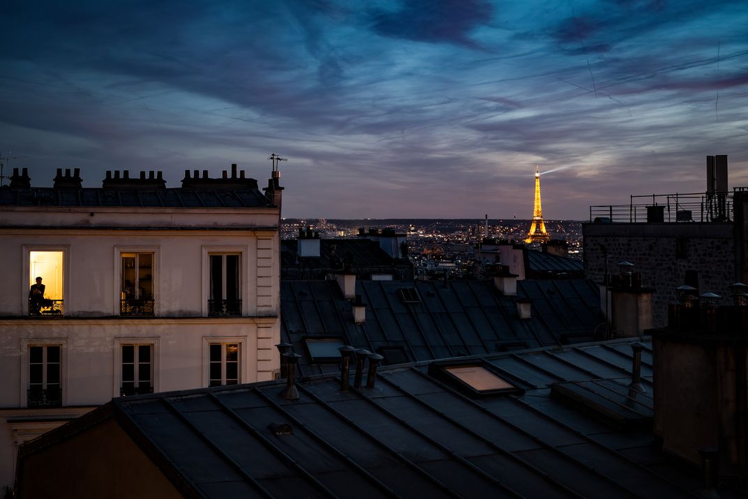 Eiffel Tower glows over Paris rooftops, a figure lit in a apartment ...